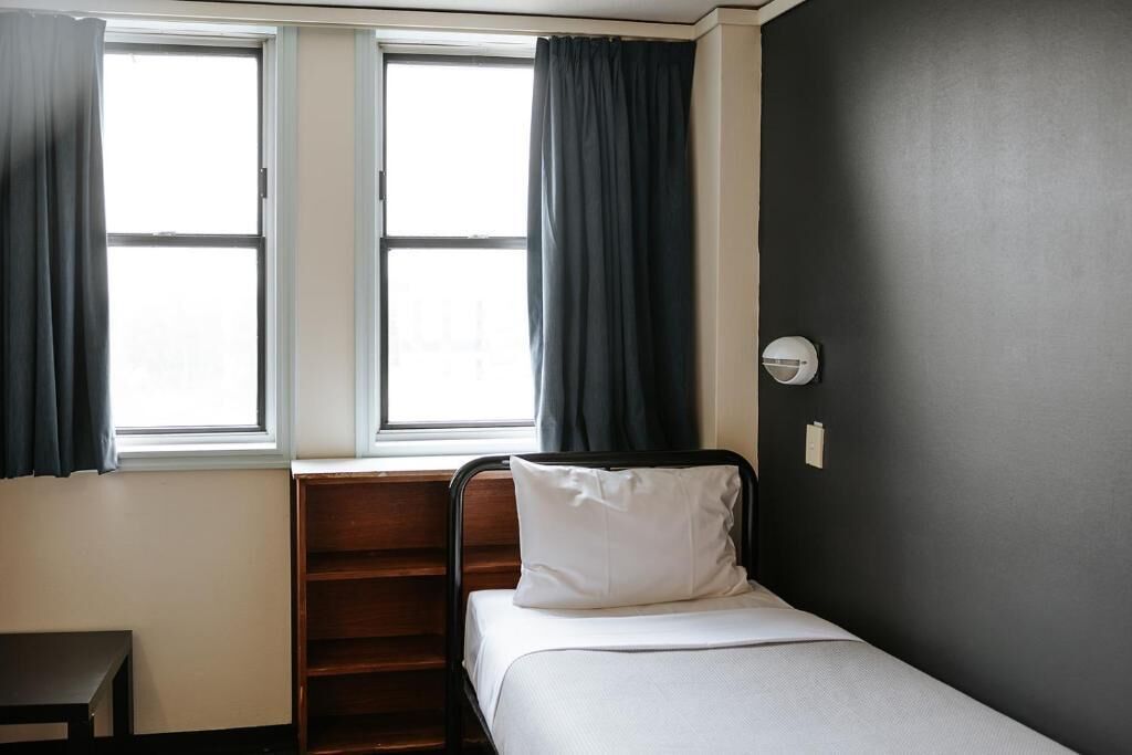 Uma cama de solteiro com colchão e travesseiro brancos, um banquinho de madeira e uma janela com cortinas.