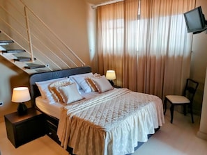 Quarto de hotel com cama, luminárias de cabeceira, uma cadeira, televisão e escada.