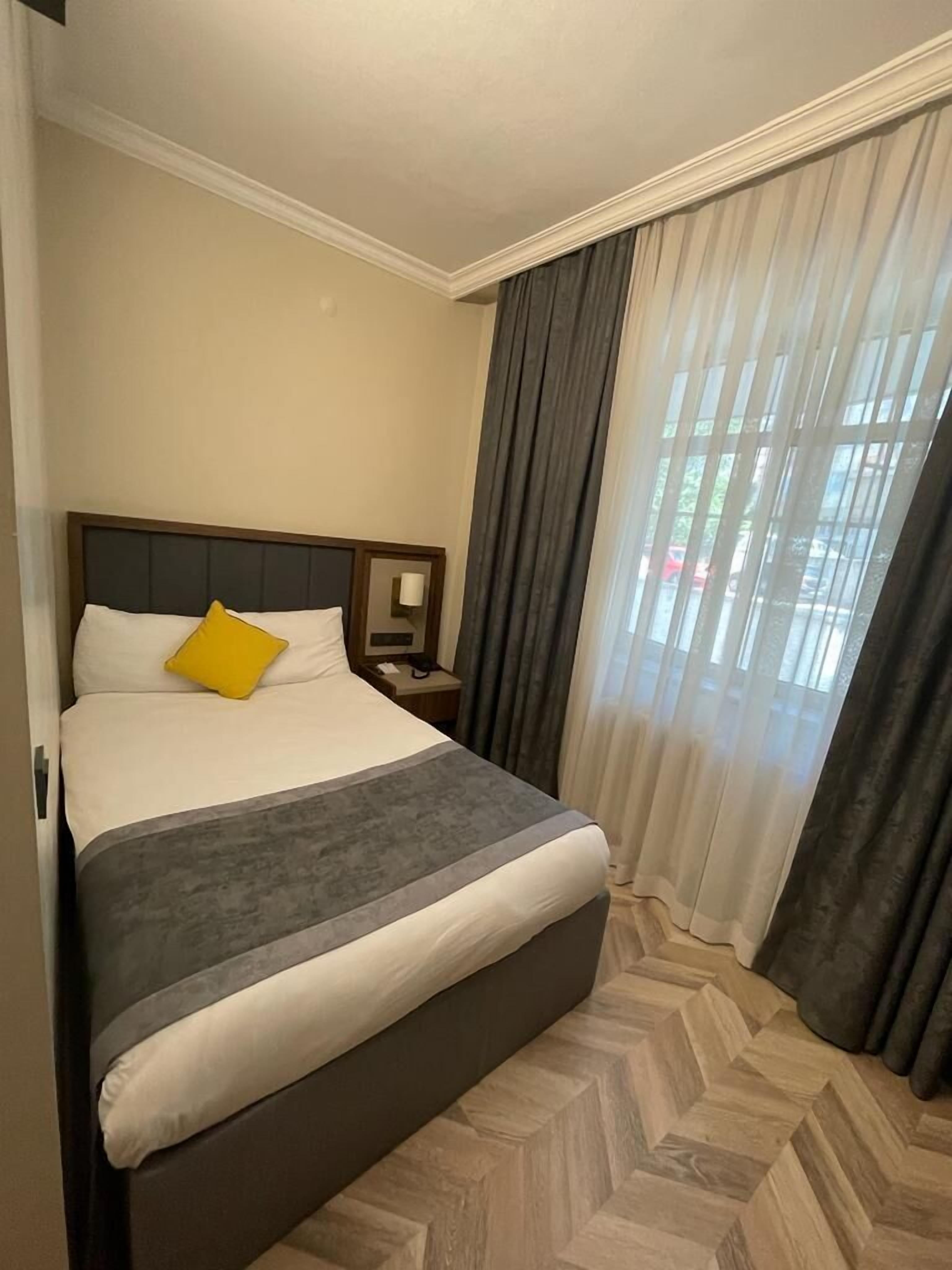 Quarto de hotel com cama, mesa de cabeceira, luminária e janela com cortinas.