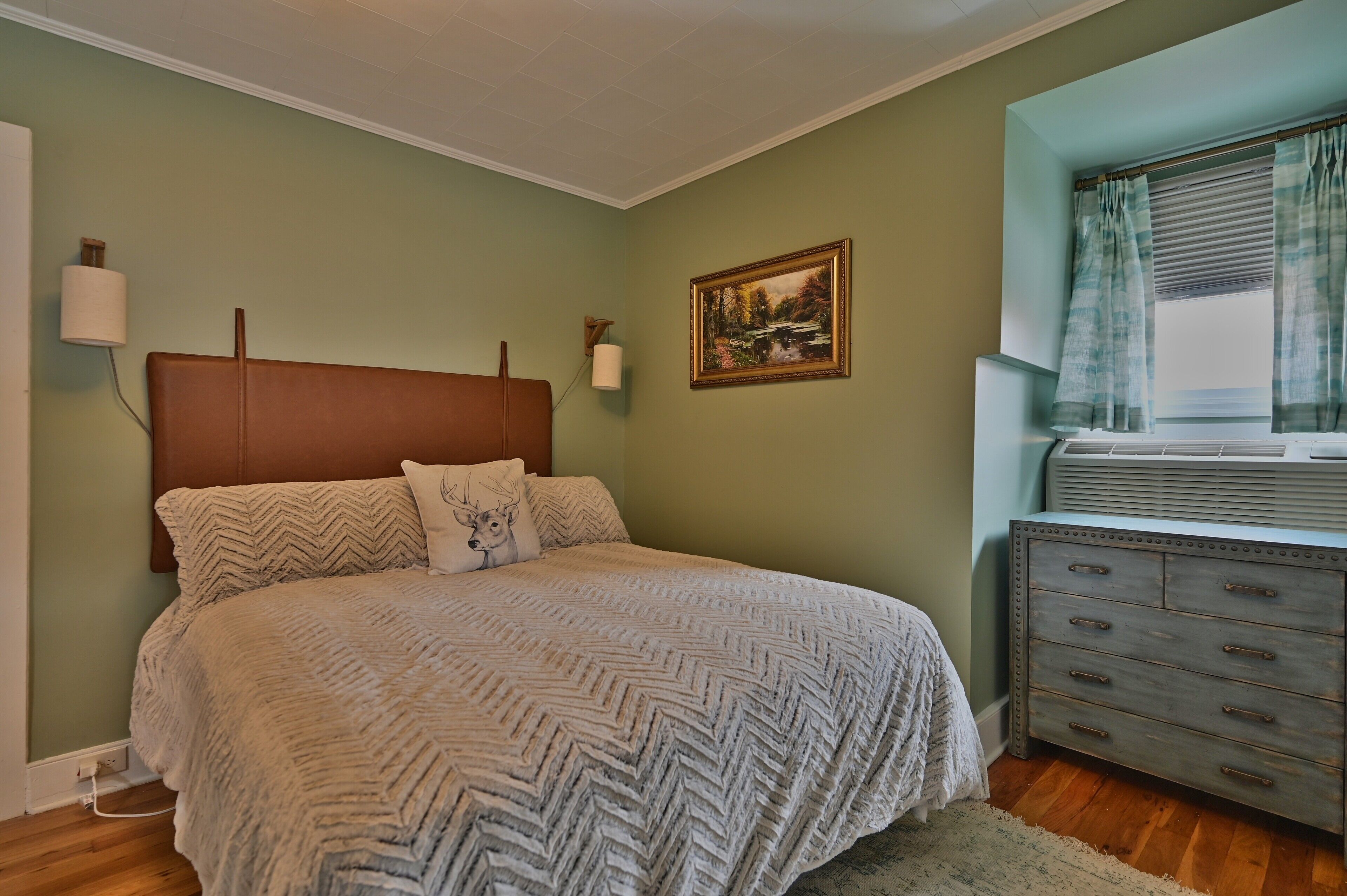 Quarto com cabeceira de madeira, cama com colcha estampada, penteadeira com gavetas, luminária fixada na parede e quadro emoldurado.