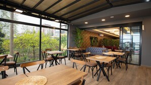 Um café moderno com mesas e cadeiras de madeira, grandes janelas com vista para árvores e edifícios, e uma área de estar acolhedora com um sofá e plantas.