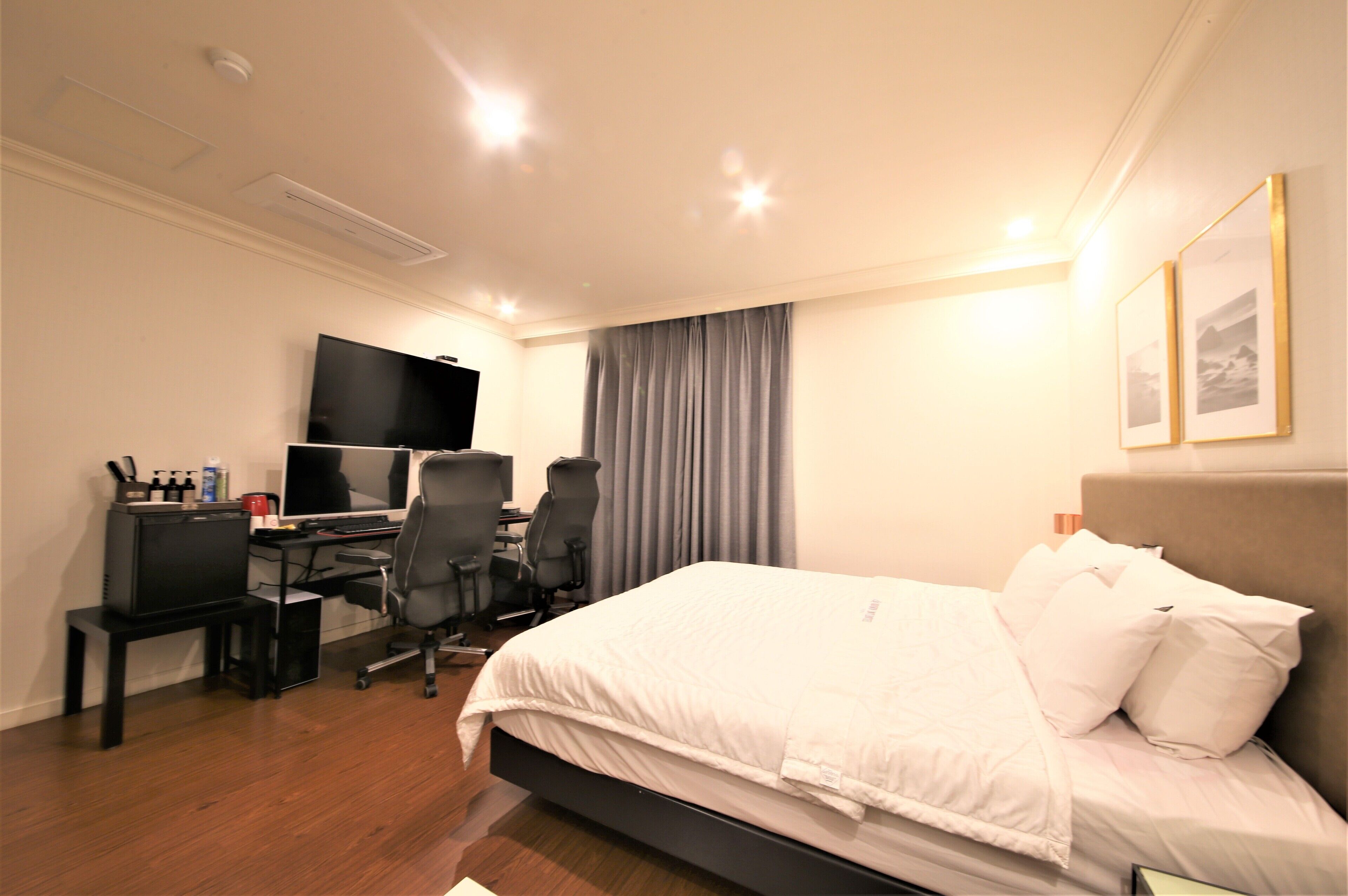 Quarto de hotel com uma cama, uma escrivaninha com computador, uma cadeira, televisão e uma janela com cortinas.