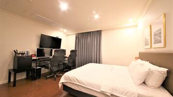 Quarto de hotel com uma cama, uma escrivaninha com computador, uma cadeira, televisão e uma janela com cortinas.