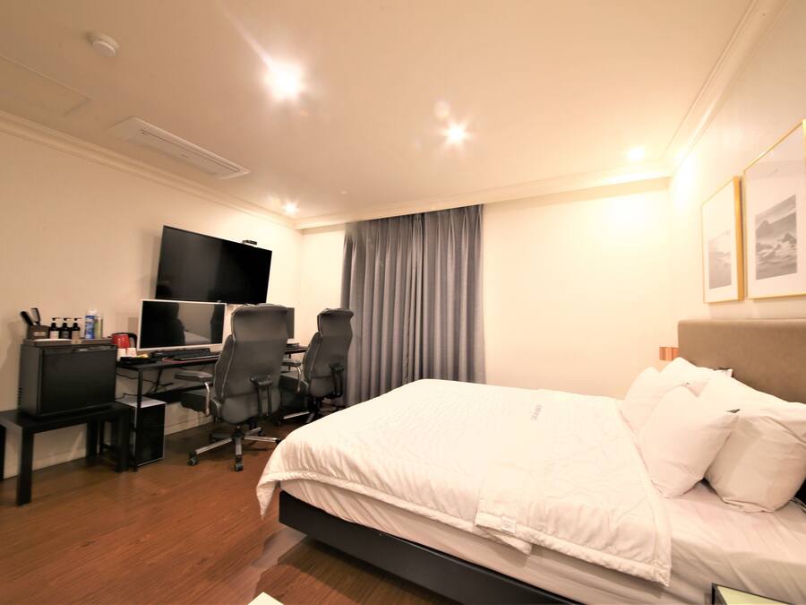 Quarto de hotel com uma cama, uma escrivaninha com computador, uma cadeira, televisão e uma janela com cortinas.