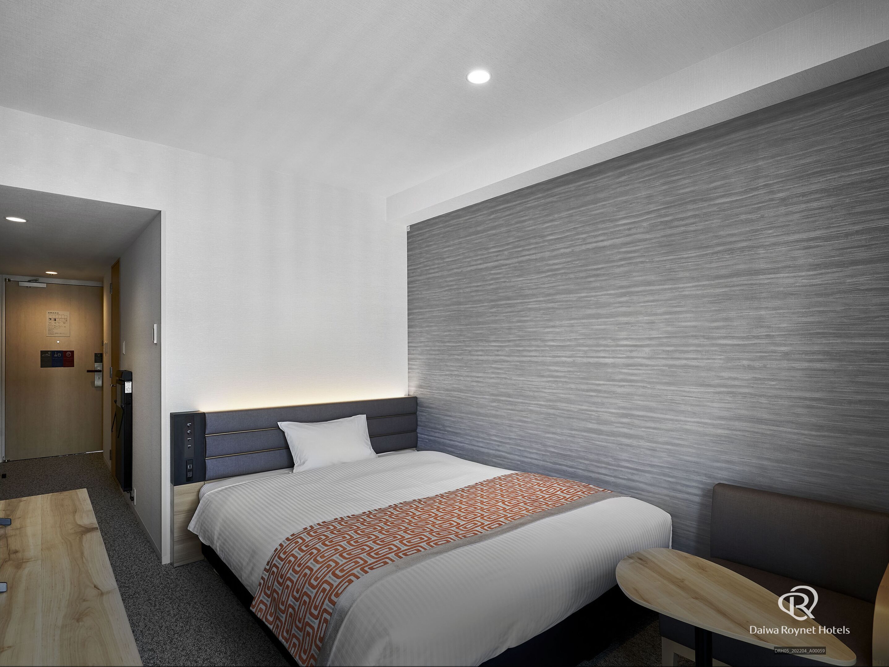 Quarto de hotel moderno com uma cama bem arrumada, uma pequena mesa de madeira e um espaço para sentar.