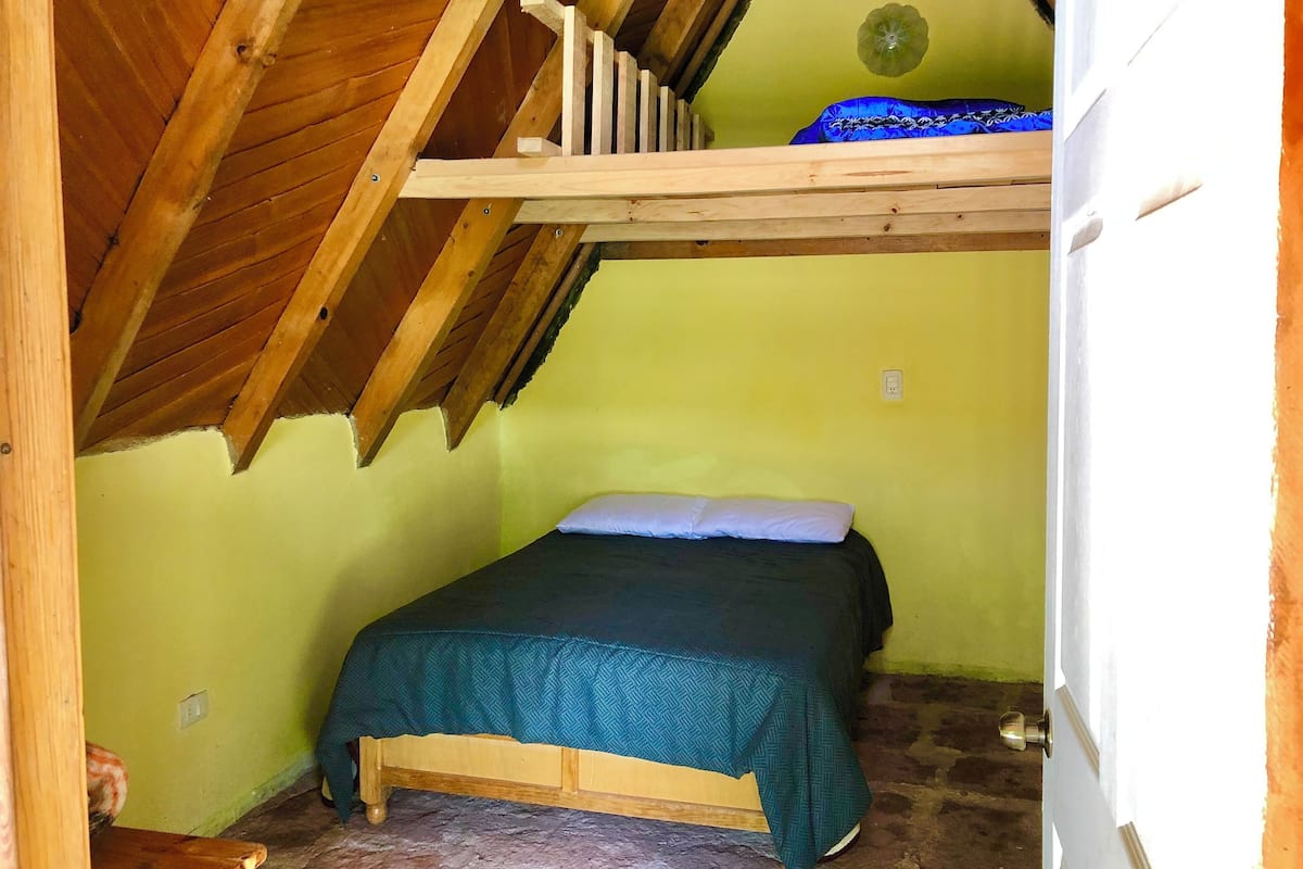 Quarto pequeno, com uma cama de solteiro, teto inclinado e vigas de madeira.