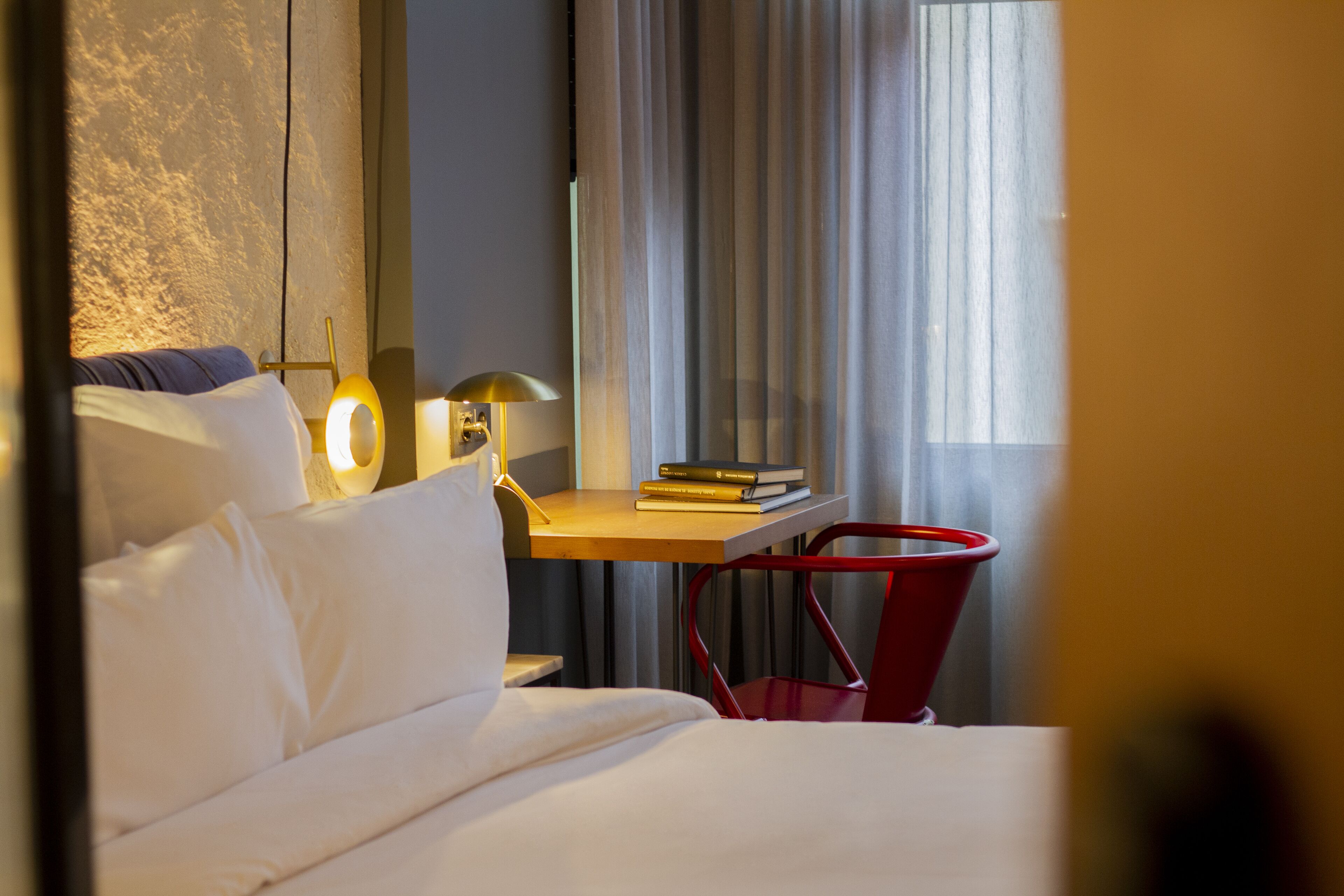Quarto de hotel com cama, escrivaninha com luminária, cadeira e janela com cortinas.