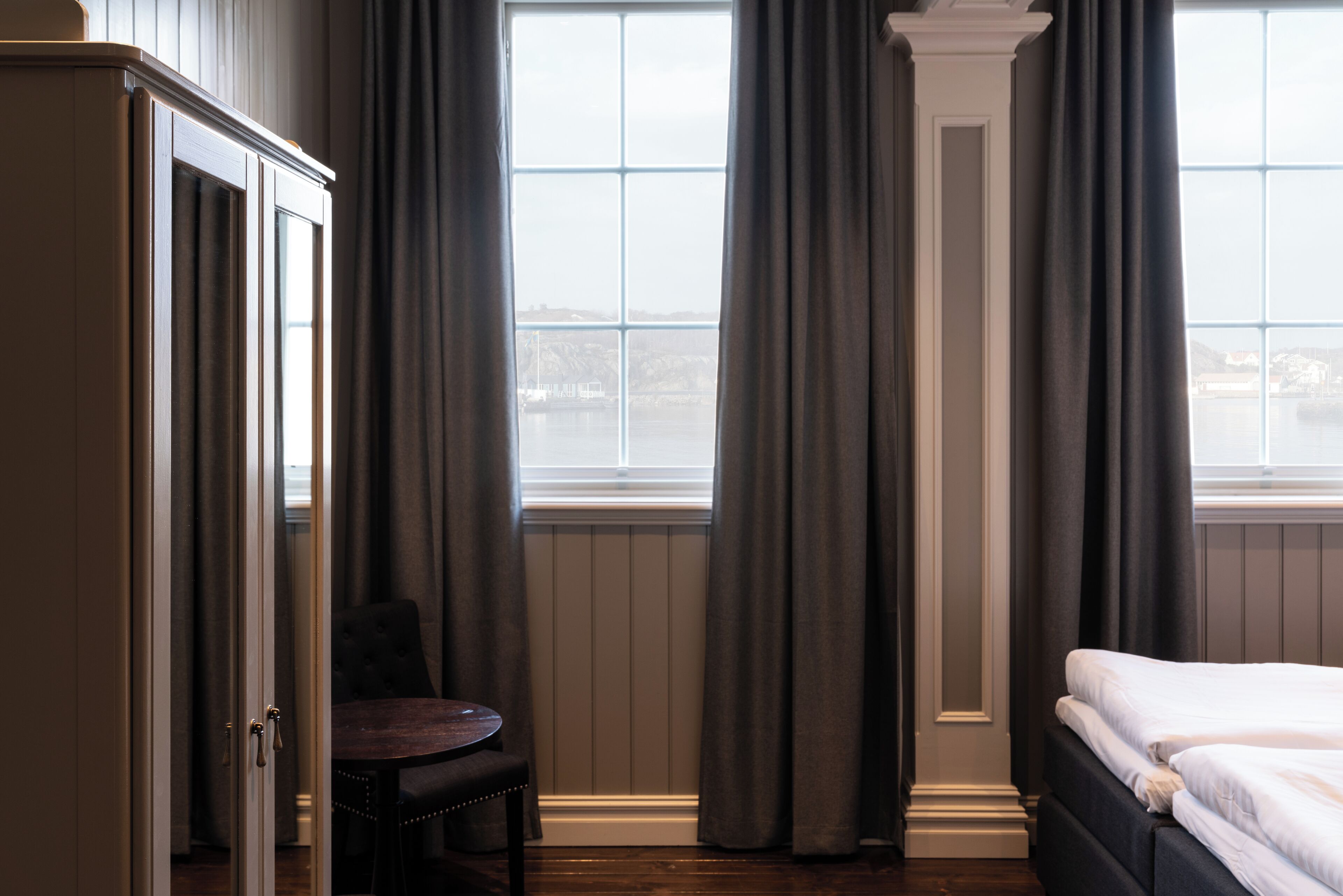 Quarto de hotel com cama, uma mesinha e uma cadeira. Há uma janela com cortinas e vista para o exterior.