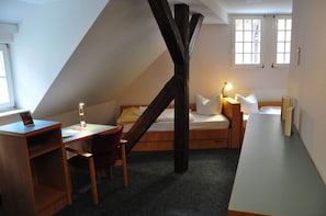 Quarto pequeno e acolhedor com teto inclinado, uma escrivaninha com cadeira, cama com almofadas e mesa de cabeceira com abajur.