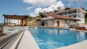 Piscina com espreguiçadeiras, pérgola e um edifício com várias andares e varandas.