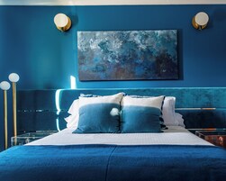 Quarto com uma cama grande, duas luminárias de cabeceira, um quadro abstrato azul e uma mesa de cabeceira com abajur.