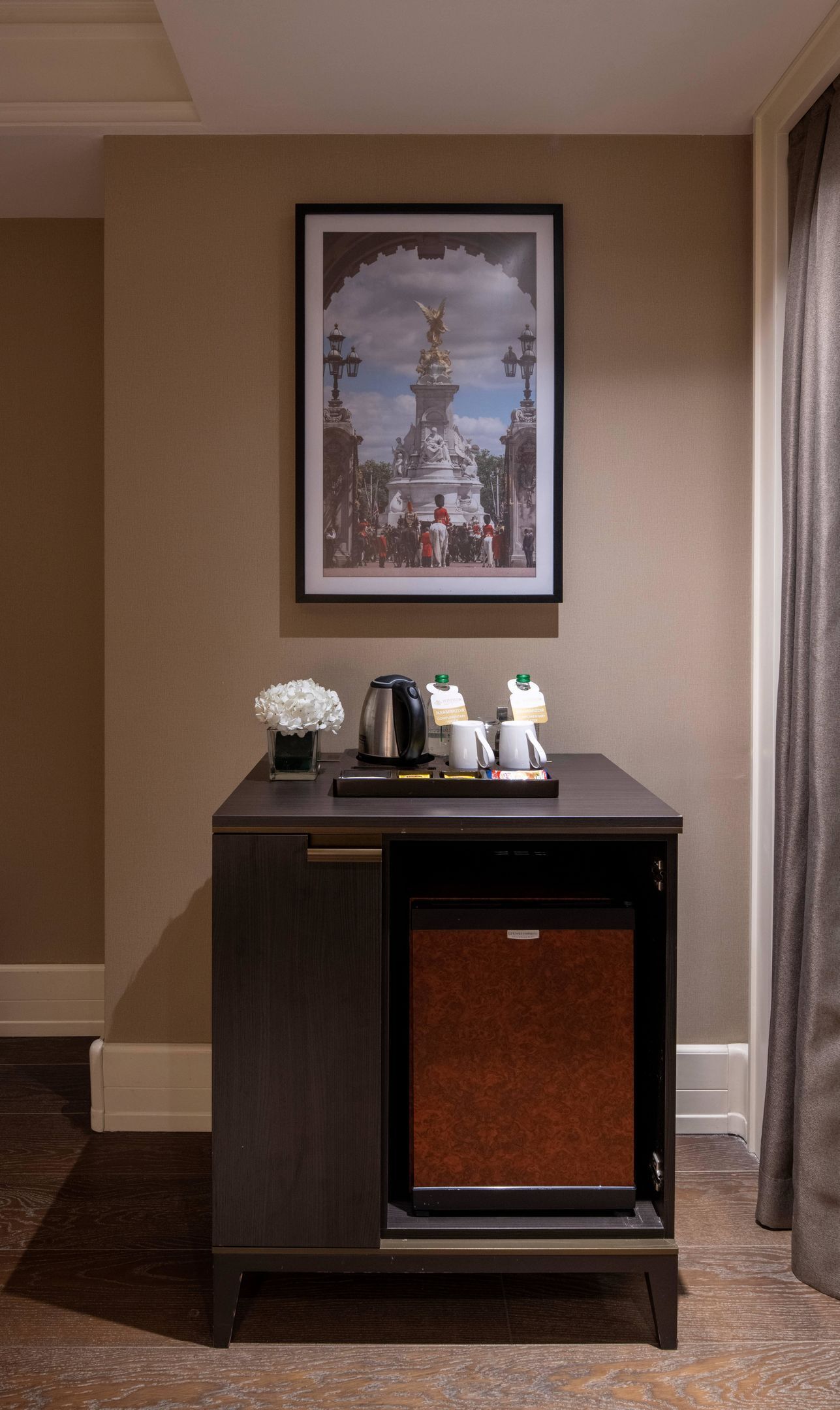 Bancada de recepção de hotel com uma fotografia histórica emoldurada acima, um pequeno vaso com flores brancas, uma cafeteira e uma bandeja com uma garrafa de água e uma caneca.