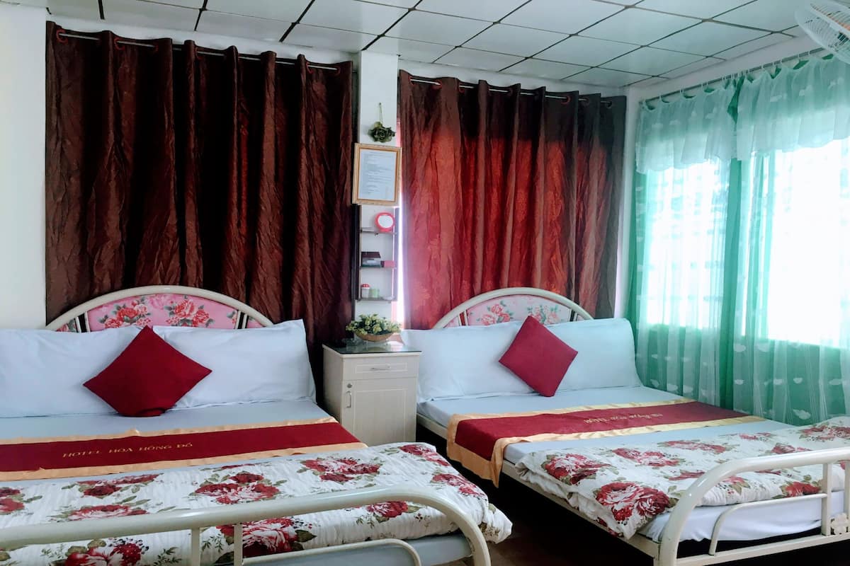 Quarto de hotel com duas camas, cada uma com roupa de cama florida e almofadas vermelhas. Há uma mesa de cabeceira com uma planta pequena e um certificado emoldurado.
