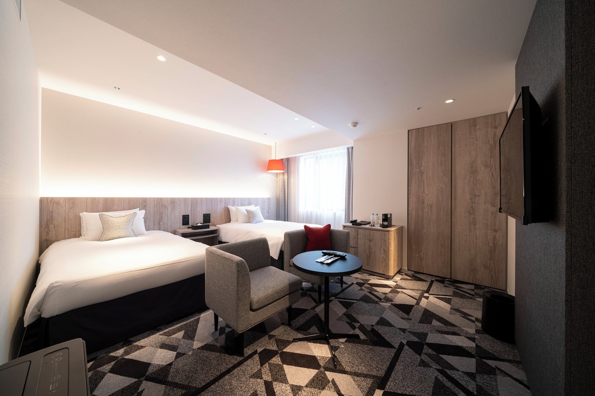 Quarto de hotel moderno com uma cama grande, um sofá, uma mesa pequena e uma televisão fixada na parede.