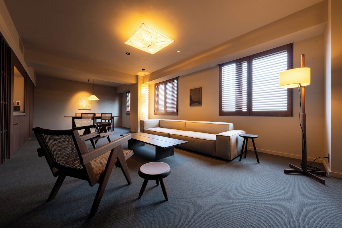 Uma sala moderna com sofá, mesa de centro e cadeiras, uma área de jantar com mesa e cadeiras, e grandes janelas com persianas.