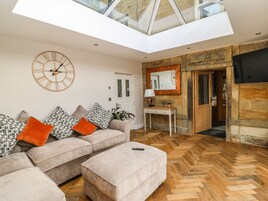 Uma sala de estar moderna com teto alto, um relógio grande, um conjunto de sofás, uma mesa consola e um chão de madeira em padrão herringbone.