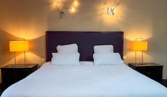 Uma cama de casal com lençóis brancos, duas mesas de cabeceira com abajures, um painel de cabeceira escuro e arandelas na parede.
