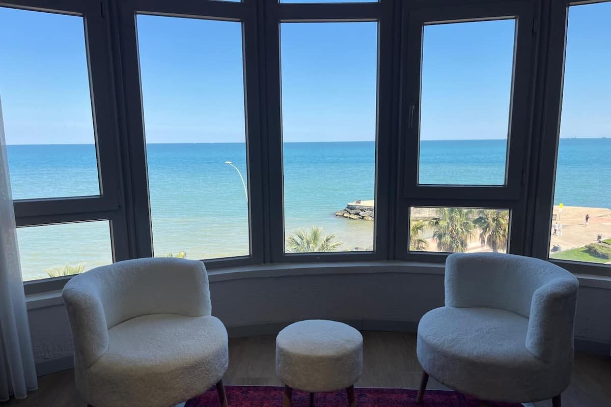 Quarto com vista para a baía, duas cadeiras brancas, um pequeno banquinho e um tapete vermelho estampado.