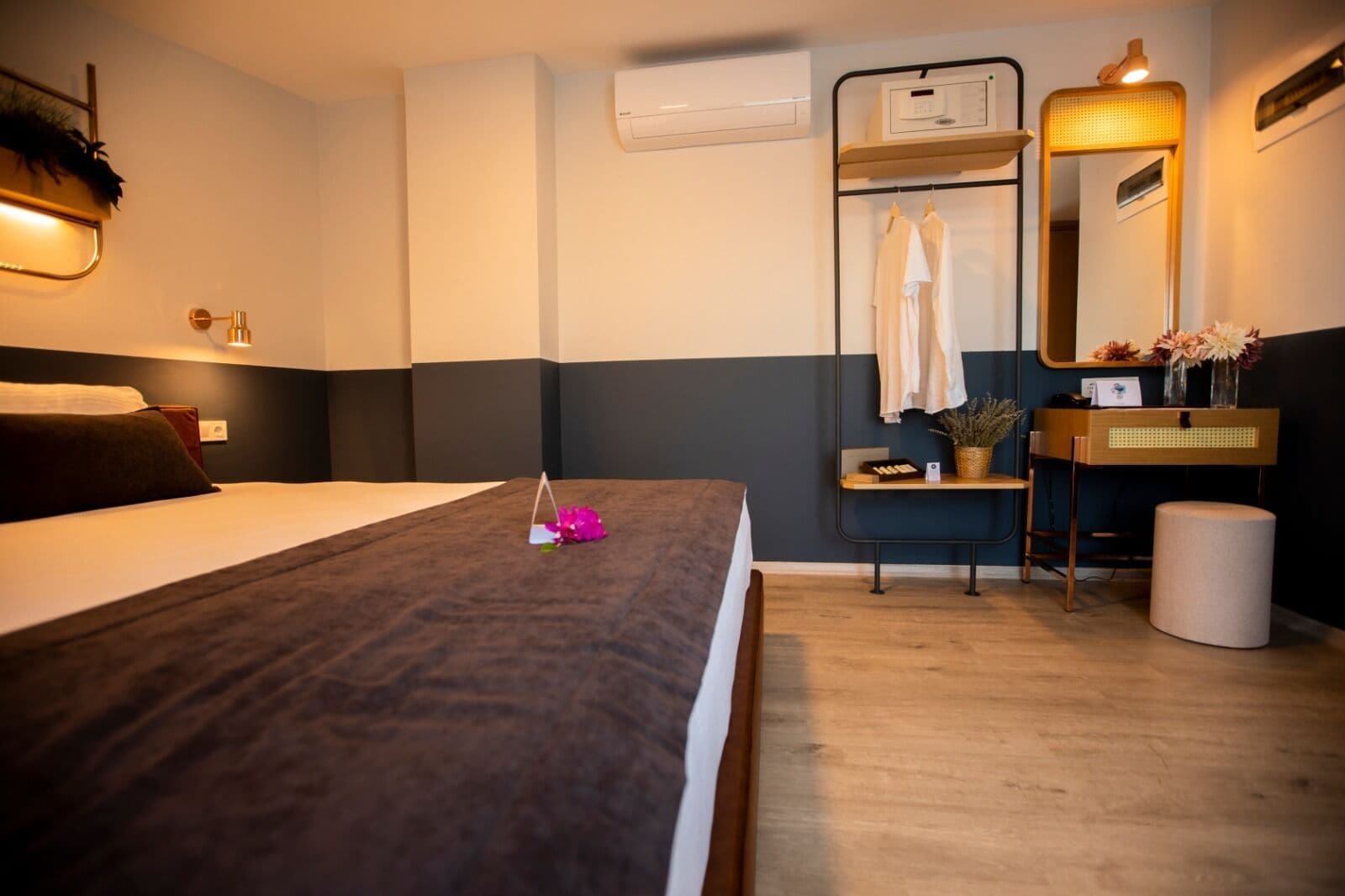 Quarto de hotel com cama, mesa de cabeceira com luminária, espelho e pequeno estante com artigos de higiene pessoal.
