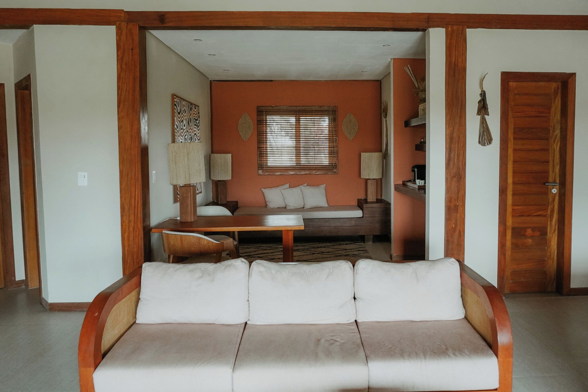 Uma sala de estar com sofá, mesa de centro e mesa de jantar. Há uma janela com persianas, um abajur e uma porta com moldura de madeira.