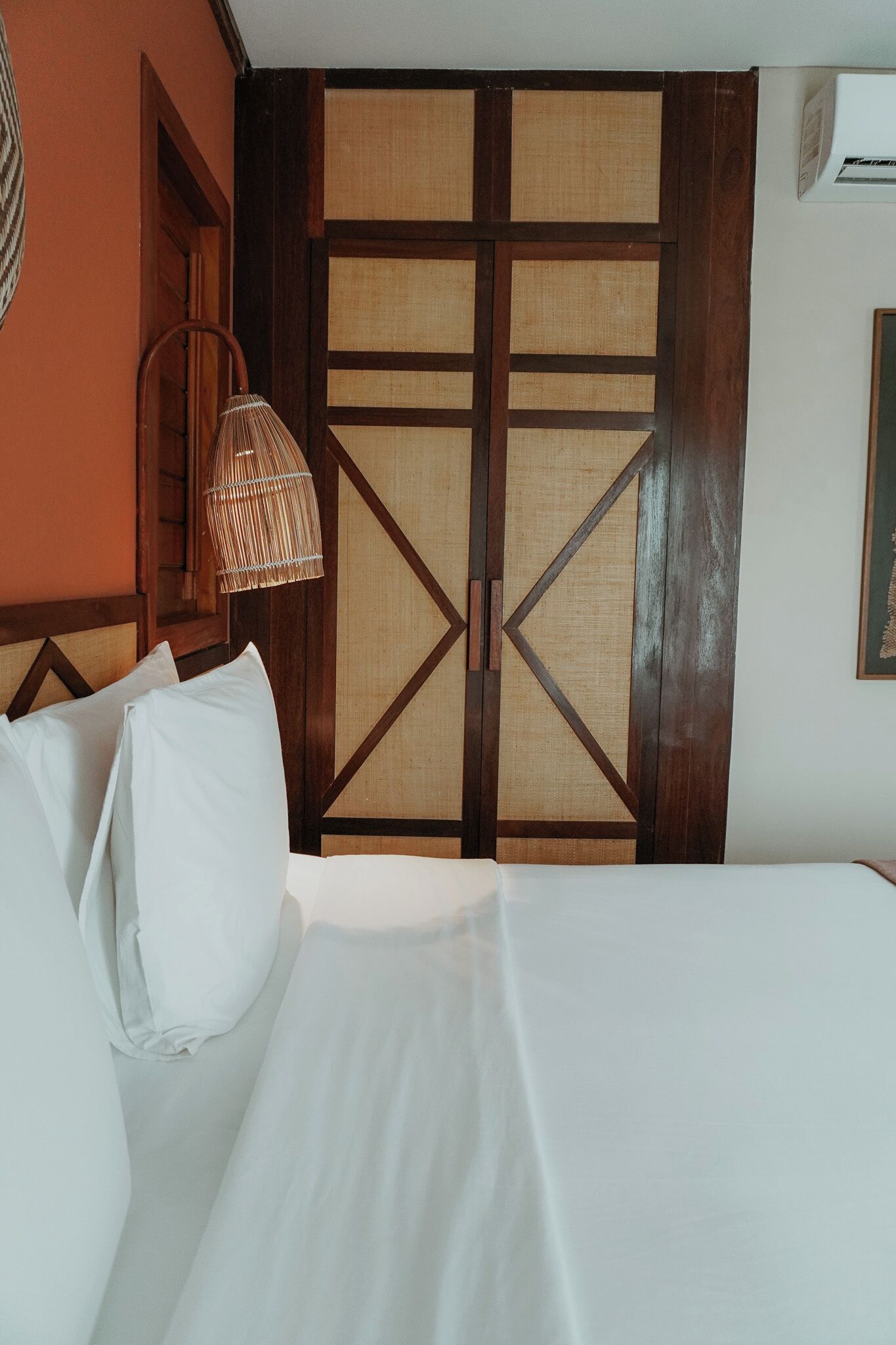 Quarto de hotel com cama, luminária de fibra natural e divisória de madeira com design geométrico.