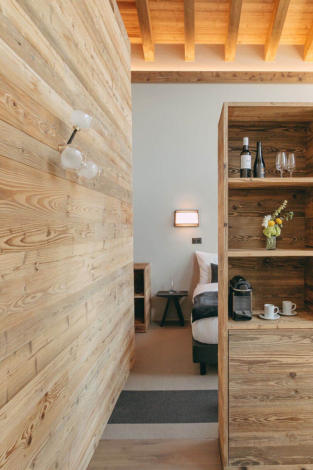 Interior em madeira com cama, mesa de cabeceira e uma estante com garrafas de vinho e copos.
