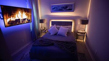 Quarto com cama, mesas de cabeceira, TV na parede exibindo uma lareira, um quadro emoldurado e uma janela com cortinas.