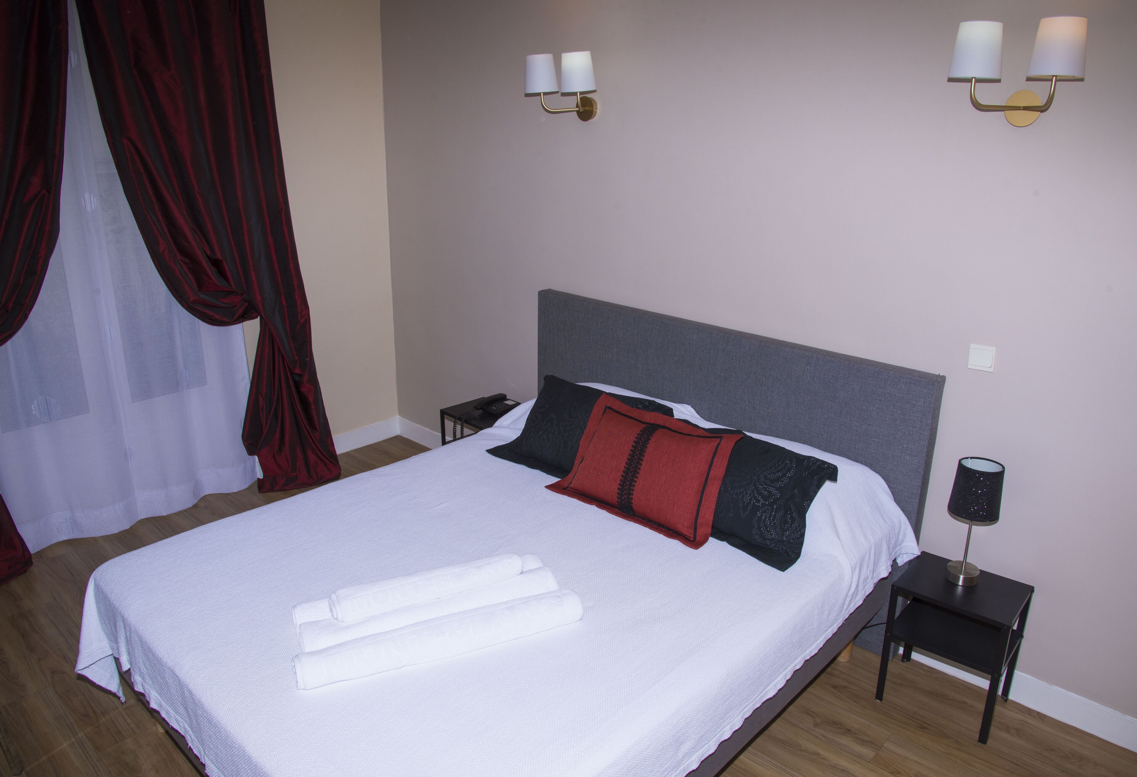 Cama bem arrumada com lençóis brancos, almofadas vermelhas e pretas e um conjunto de toalhas brancas. Existem duas mesas de cabeceira, uma com abajur e a outra com telefone. O quarto tem piso de madeira e uma parede lisa com duas luminárias de parede.