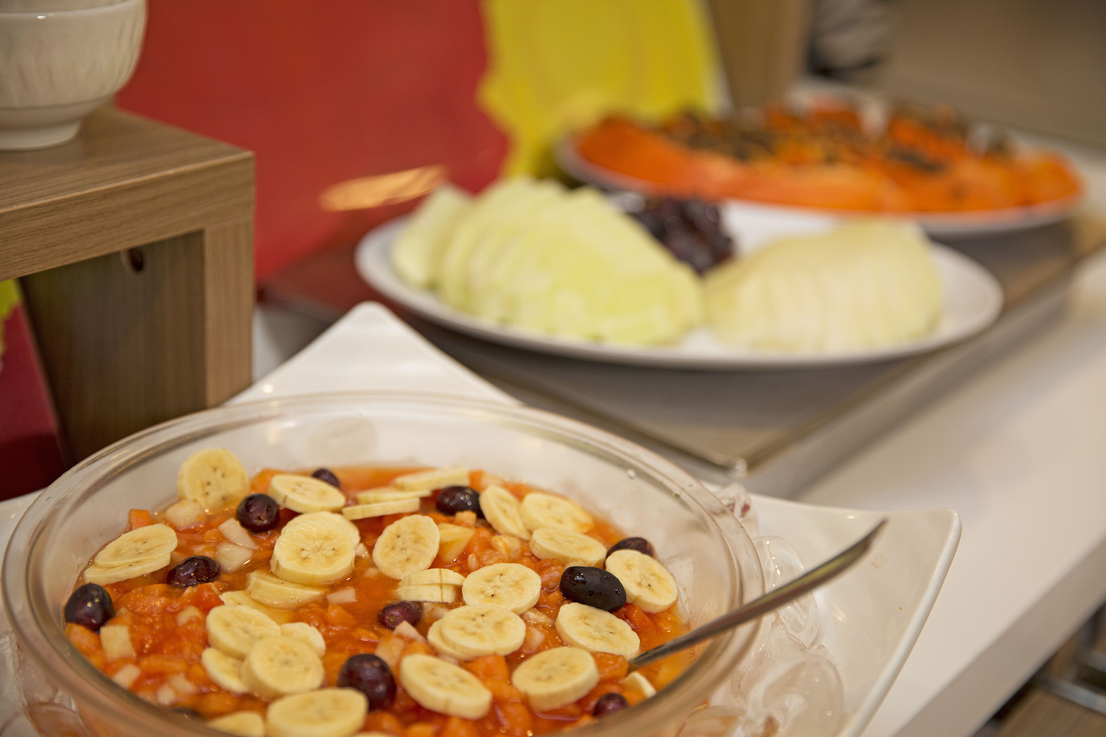 Buffet de café da manhã com diversos pratos, incluindo uma tigela de cereal coberta com bananas e passas, e pratos com frutas fatiadas.