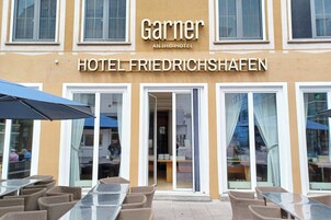 Exterior do Hotel Friedrichshafen, com entrada desobstruída, área de estar ao ar livre e interior visível através das portas de vidro.