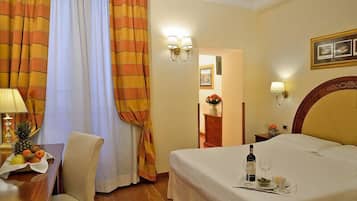 Quarto de hotel com uma cama, uma escrivaninha com uma cesta de frutas, uma luminária e uma janela com cortinas.