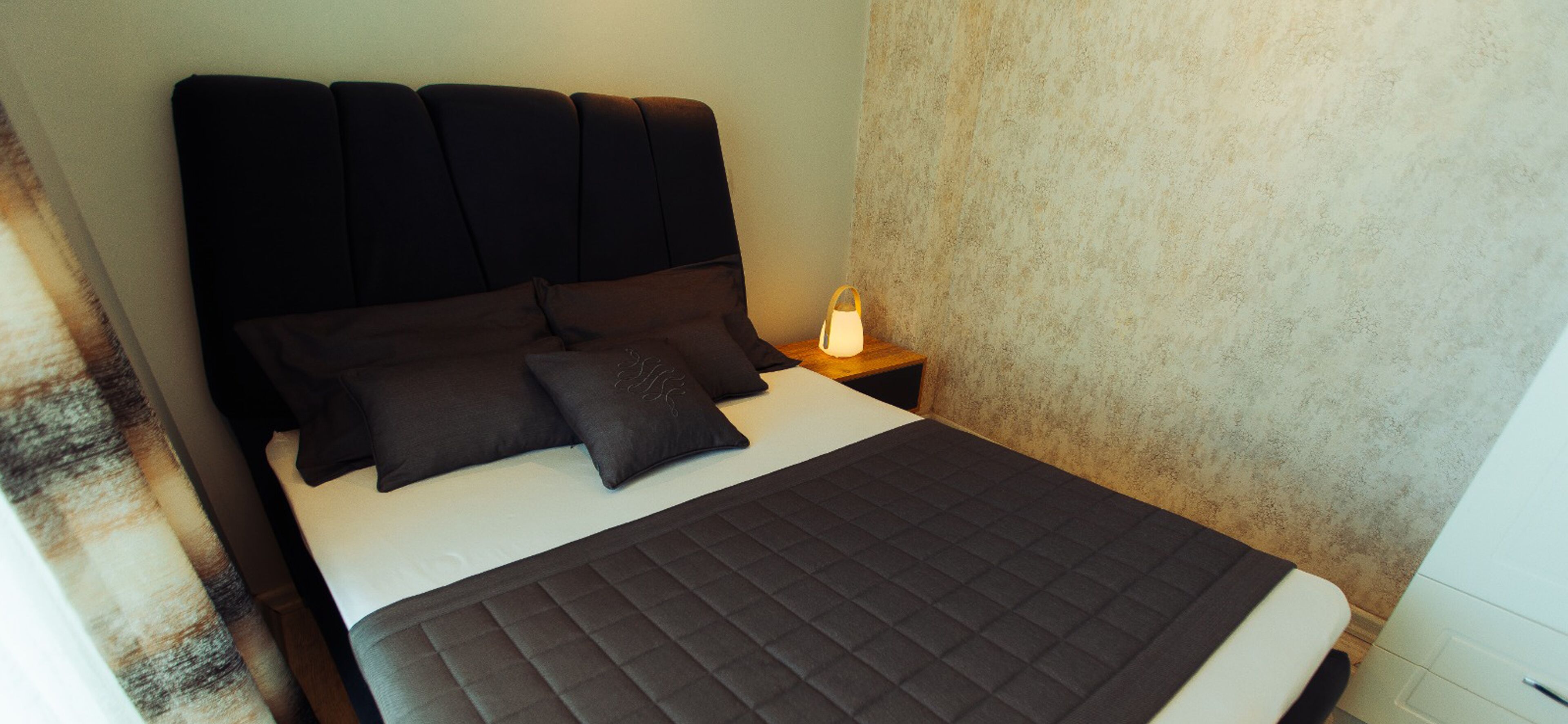 Uma cama pequena e bem arrumada, com cabeceira preta e almofadas, mesa de cabeceira com abajur e parede com textura.