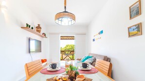 Uma sala de jantar com uma mesa de vidro, individuais de vime e um objeto decorativo central. Há duas cadeiras de madeira, um sofá e uma televisão fixada na parede.