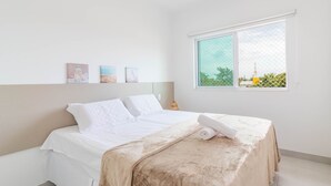 Uma cama bem arrumada com lençóis brancos e um cobertor bege, uma pequena luminária de cabeceira e três quadros nas paredes.