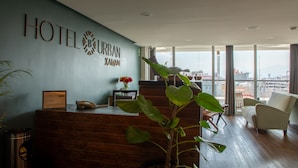 Uma área de recepção de hotel com uma mesa de madeira, uma planta e vista para a cidade através de grandes janelas.