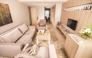 Quarto de hotel moderno com sofá, poltrona, mesa de centro, televisão e uma pequena escrivaninha com um vaso de flores.