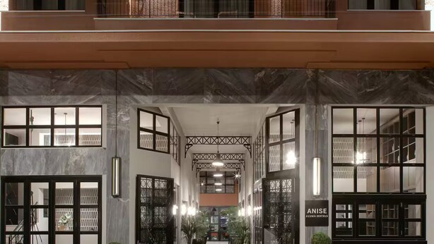 Entrada de hotel com fachada imponente, grandes portas de vidro e corredor central que leva ao interior.