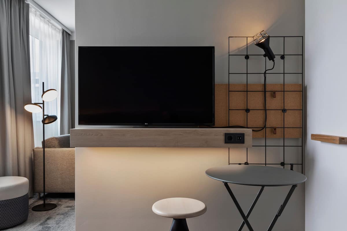 Quarto moderno com TV de tela plana fixada em um painel de madeira, uma pequena mesa redonda, um banquinho e um abajur.