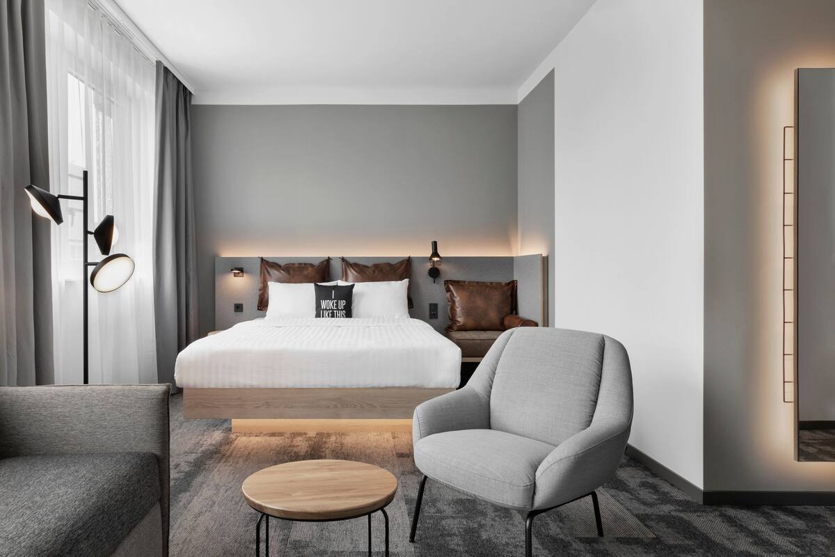 Quarto de hotel moderno com uma cama grande, uma poltrona cinza, uma pequena mesa redonda e uma luminária fixada na parede.