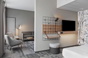 Quarto de hotel moderno com um sofá cinzento, uma pequena mesa redonda, uma poltrona cinzenta, uma TV fixada numa parede de grade e um papel de parede estampado.