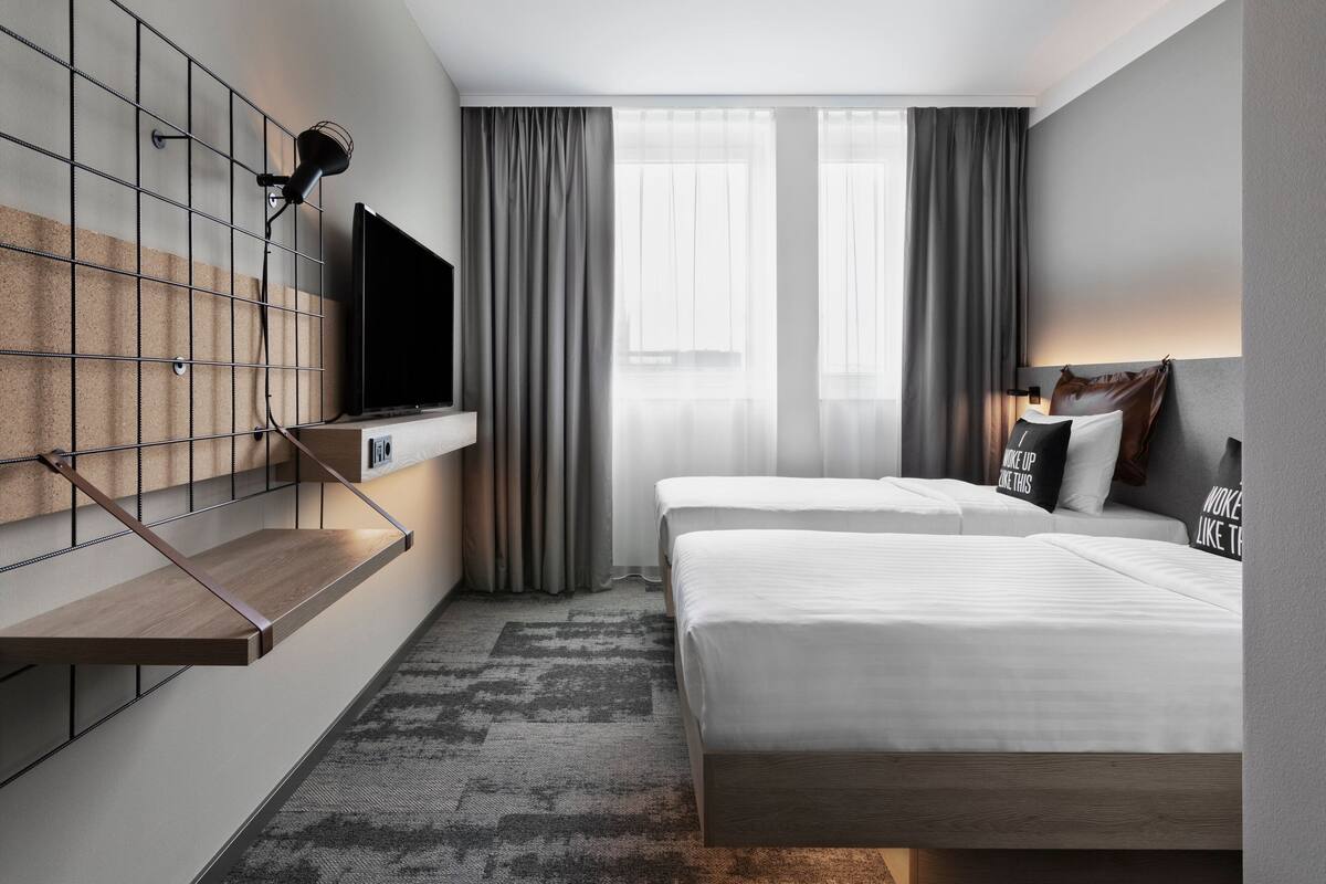Quarto de hotel com duas camas, uma televisão fixada na parede, uma estante e uma janela com cortinas.