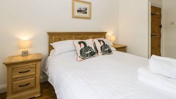 Cama bem arrumada com roupa de cama branca e duas almofadas decorativas com cisnes pretos. Há duas mesas de cabeceira com abajures, uma cabeceira de madeira e um quadro na parede.