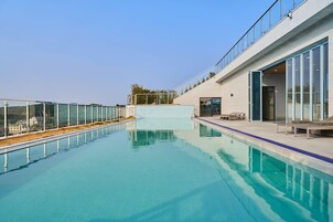Uma área de piscina moderna com água azul cristalina, guarda-corpo de vidro e um edifício com janelas amplas e porta.