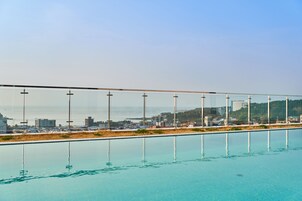 Piscina com barreira de vidro e vista para edifícios e vegetação ao fundo.
