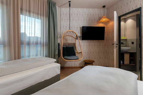 Quarto de hotel com duas camas, uma cadeira suspensa, televisão e banheiro.