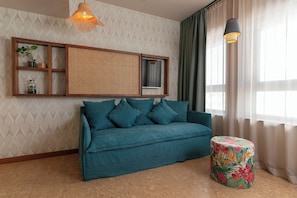 Um sofá azul-turquesa com almofadas, uma estante de prateleiras trançadas, um banquinho redondo pequeno com capa floral e duas luminárias pendentes.