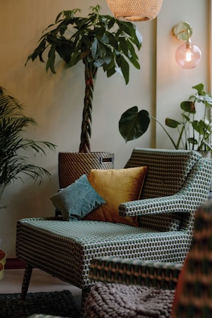 Uma cadeira de vime com almofada, um abajur e um vaso de planta num quarto.