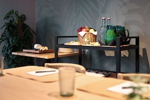 Uma área de refeições com mesa posta, uma planta e um carrinho de buffet com comida e bebidas.
