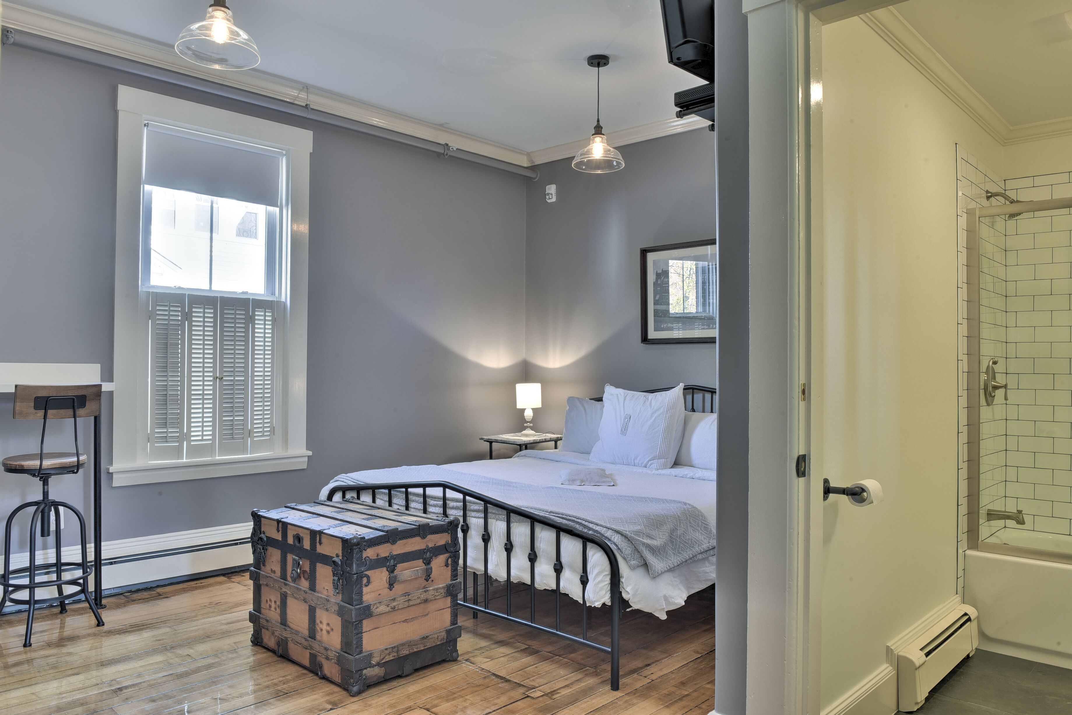 Quarto com cama, penteadeira, janela com persianas, luminária pendente e porta para um banheiro.