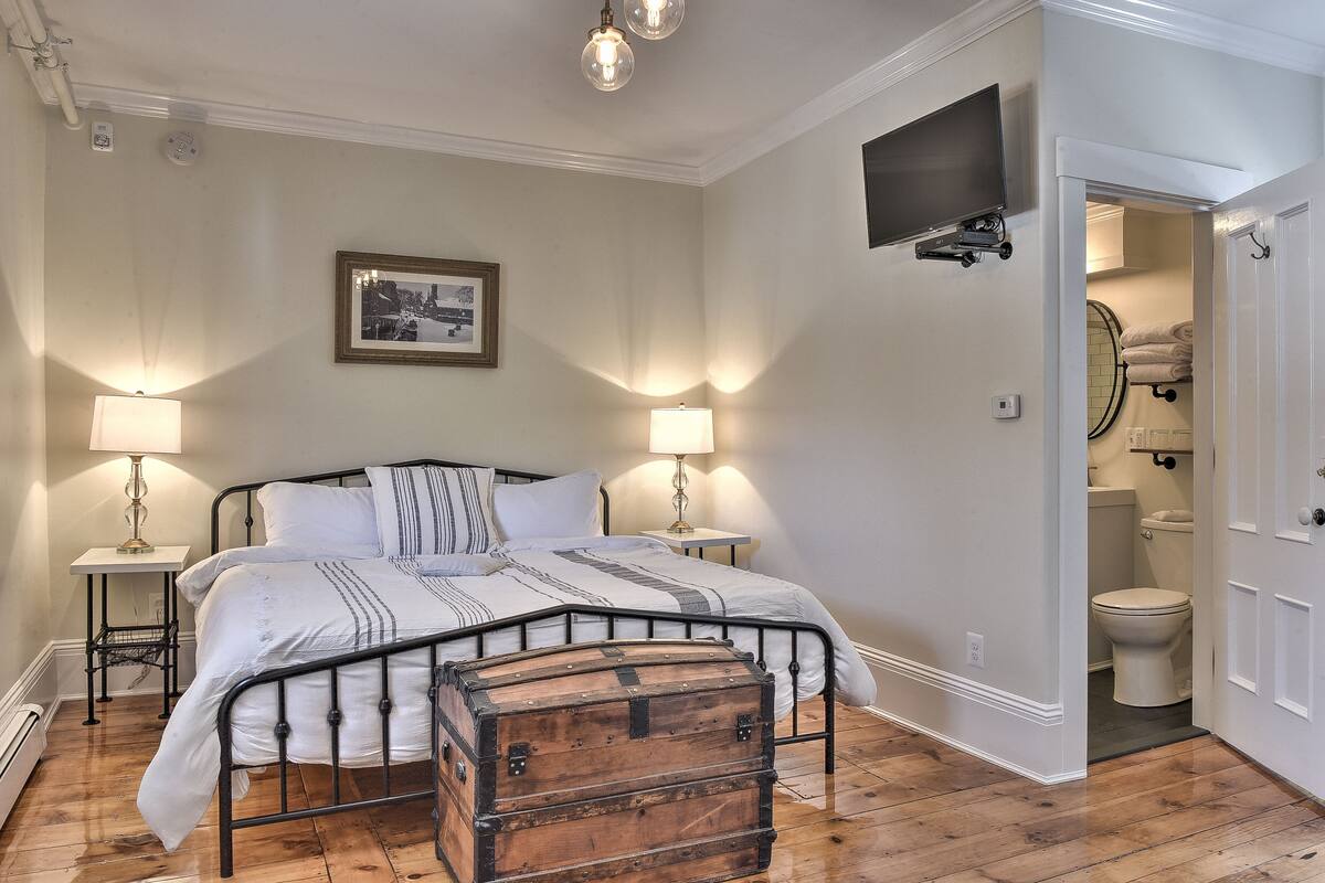 Quarto com cama de madeira, duas luminárias de apoio, televisão montada, quadro emoldurado na parede e um baú de madeira.
